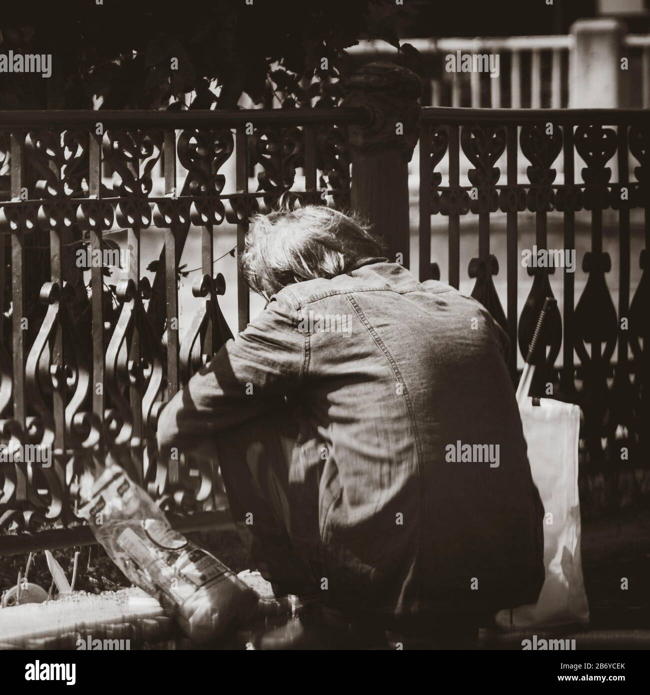 Homeless man crouching near iron fence with plastic bags around him ...