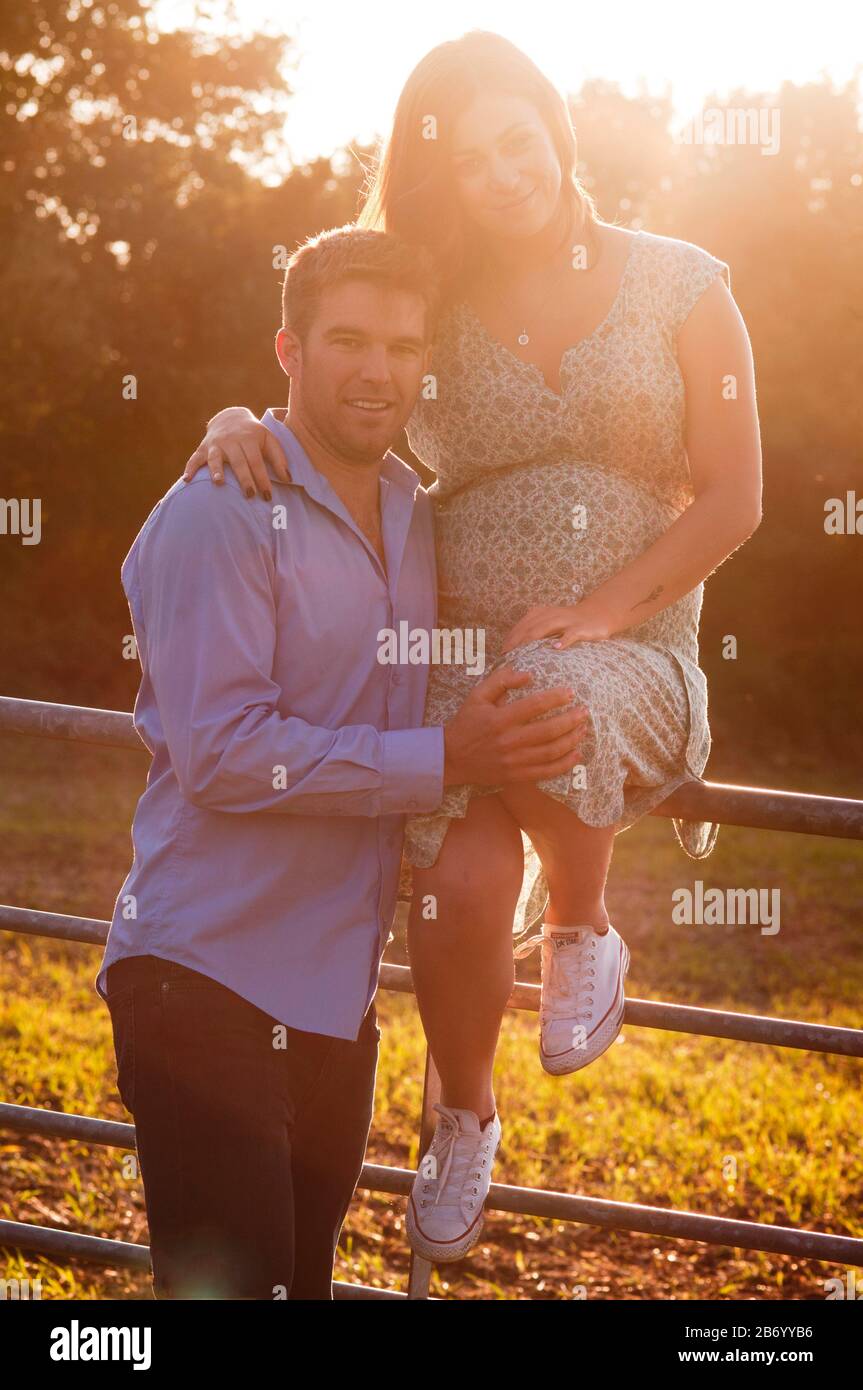 Pregnant young woman sitting on a gate with her arms around her partner ...