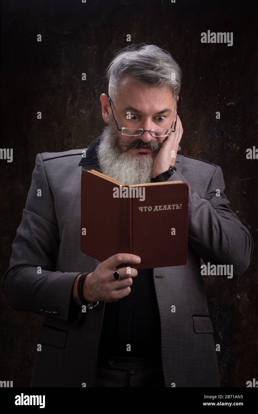 Portrait of a gray-haired bearded man with glasses reading book in ...