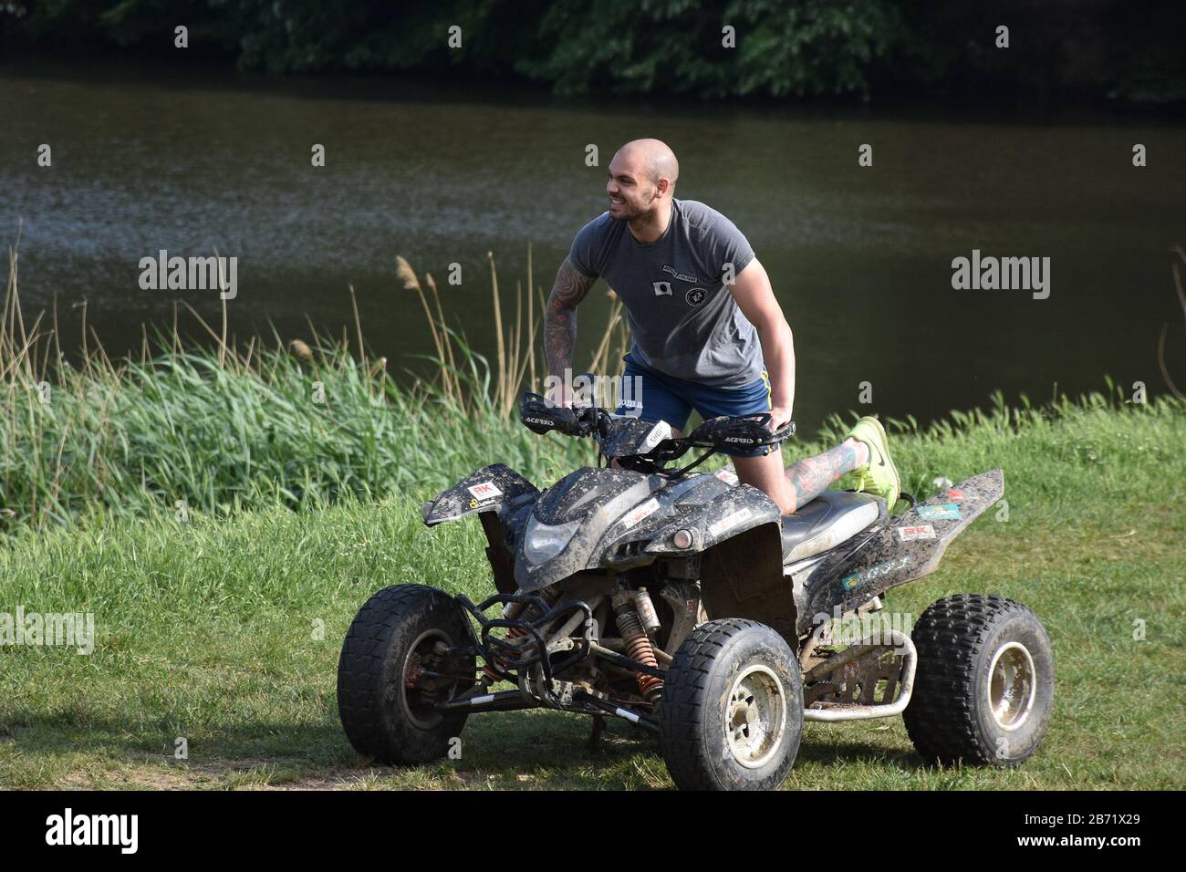 A man in a standing stance drives a quad along the grass by the river ...