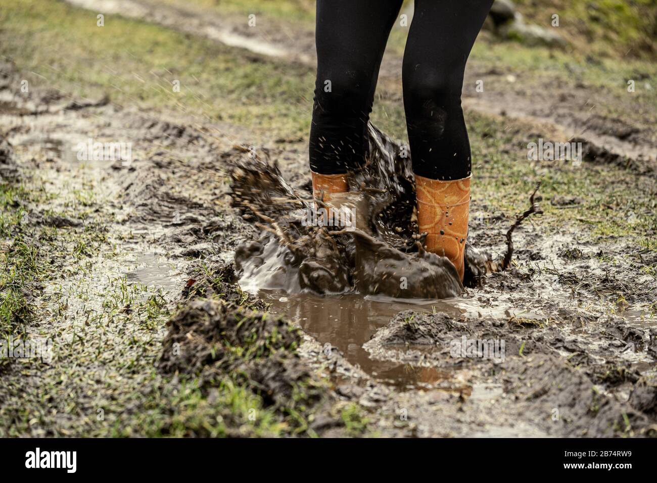 woman jumping in the puddle of mud in autumn with orange rubber boots ...