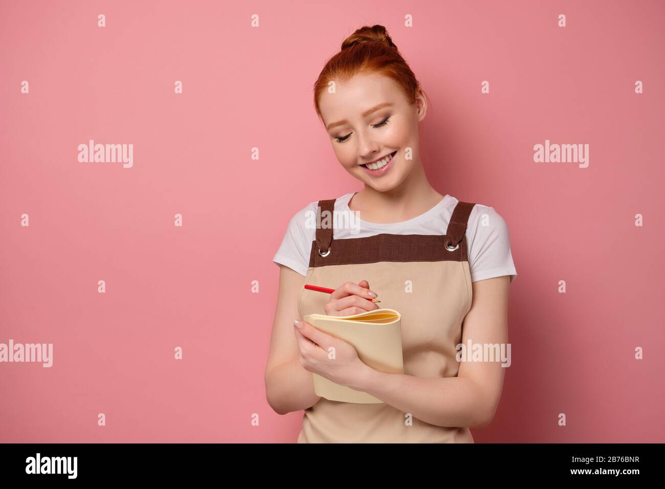 Red-haired girl in an apron with collected hair is standing on a pink ...
