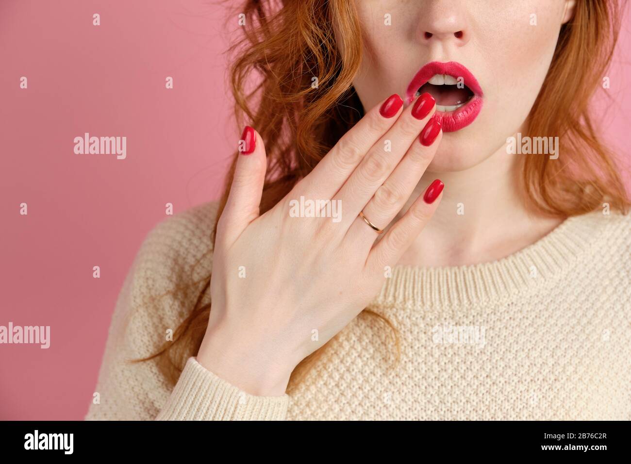 A close shot of a red-haired girl with red manicure and lipstick, covering hand with a surprised ...