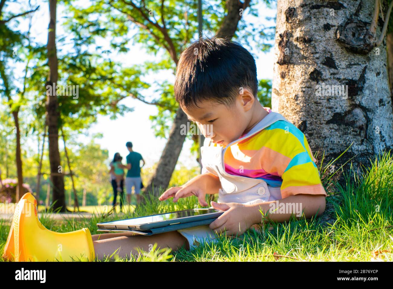 Adorable asian boy sitting under tree use tablet computer technology ...