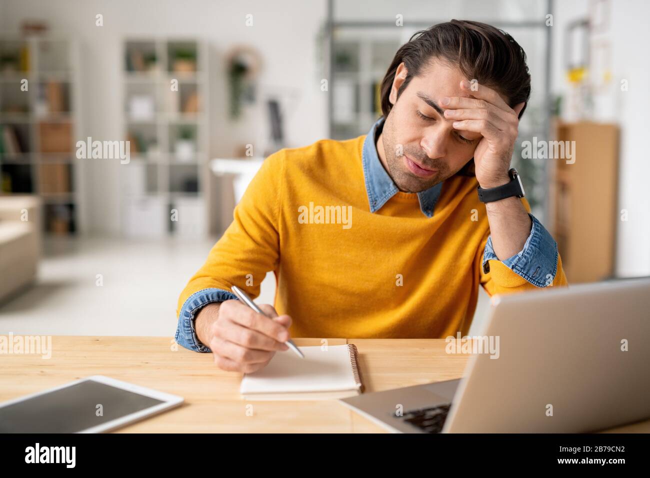 Young pensive businessman with pen sitting by desk and bending over ...