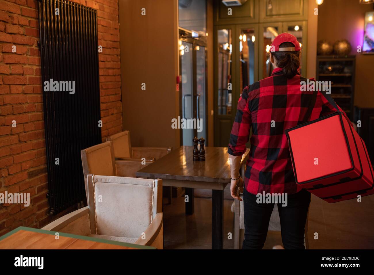 Rear view of young delivery service courier in flannel carrying big red ...