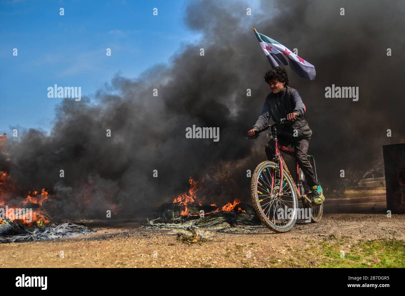 Idlib, Syria. 15th Mar, 2020. A picture shows a Syrian boy riding his bike and carrying the flag ...