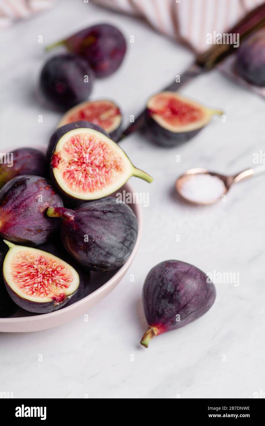 A bowl of whole and halved purple figs on a white marble countertop ...