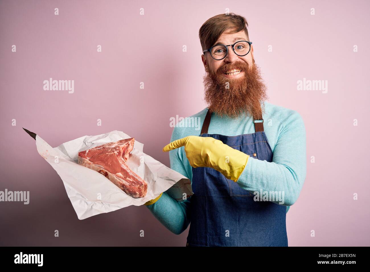 Redhead Irish butcher man with beard holding raw beef steak over pink ...