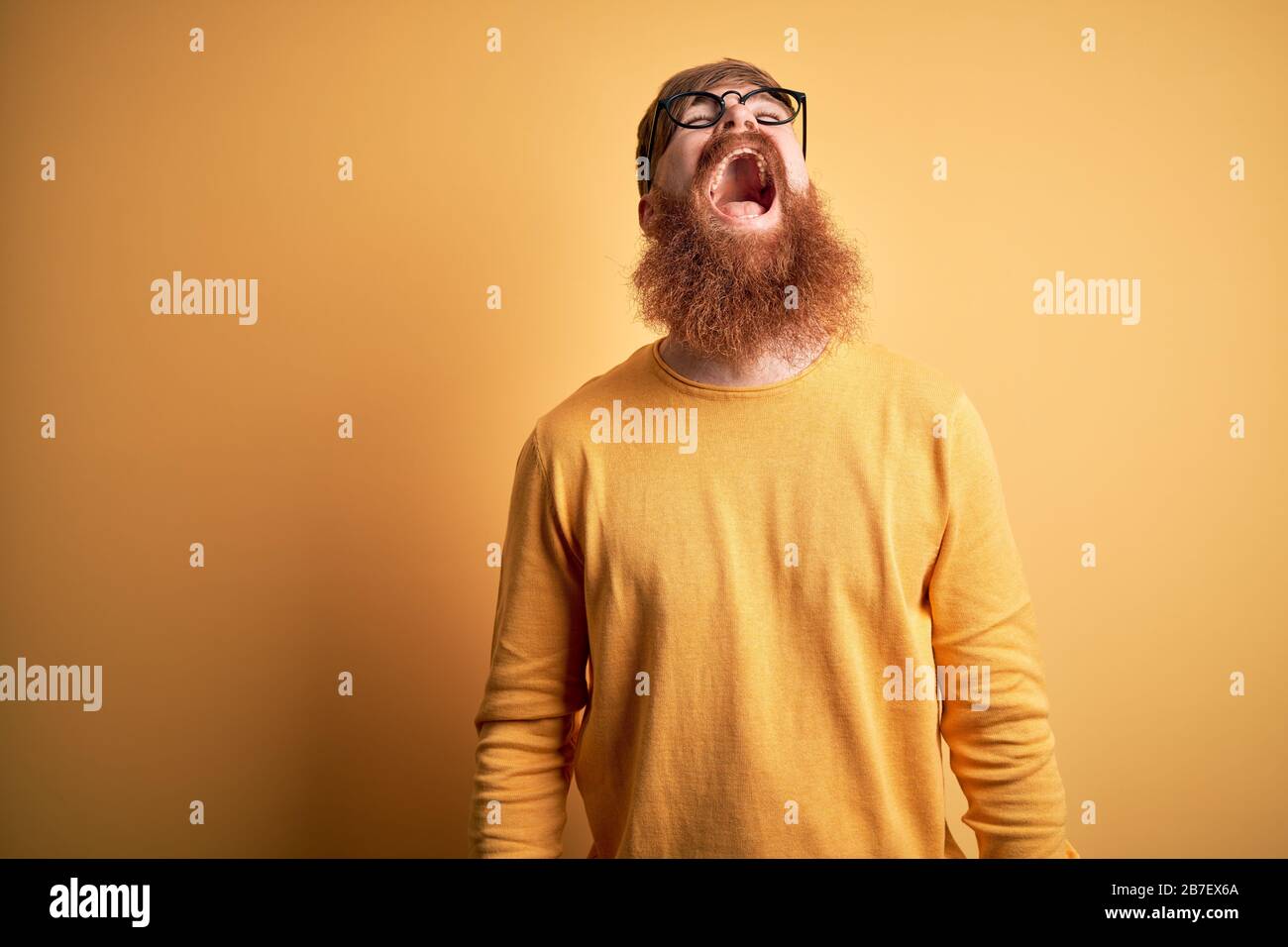 Handsome Irish redhead man with beard wearing glasses over yellow ...