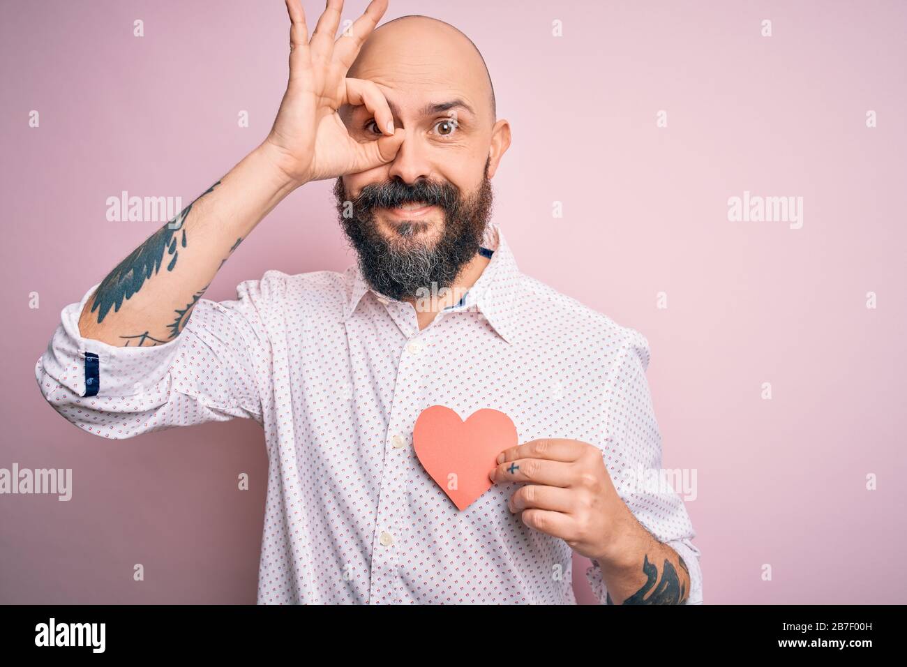 Handsome romantic bald man with beard holding red heart paper over pink ...