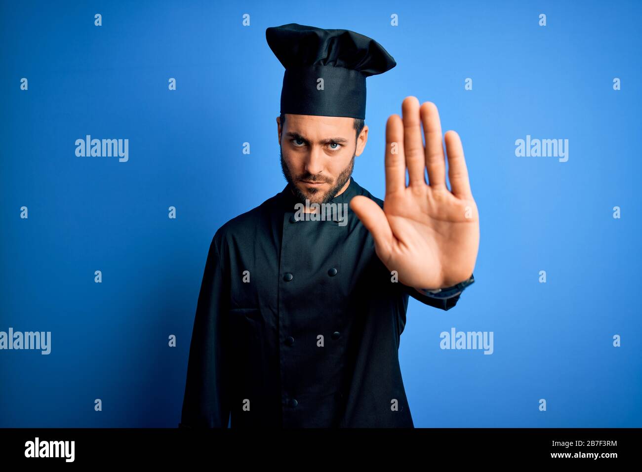 Young handsome chef man with beard wearing cooker uniform and hat over ...