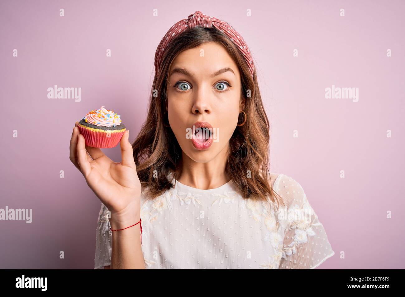 Young blonde girl eating a sweet chocolate cupcake over pink isolated ...