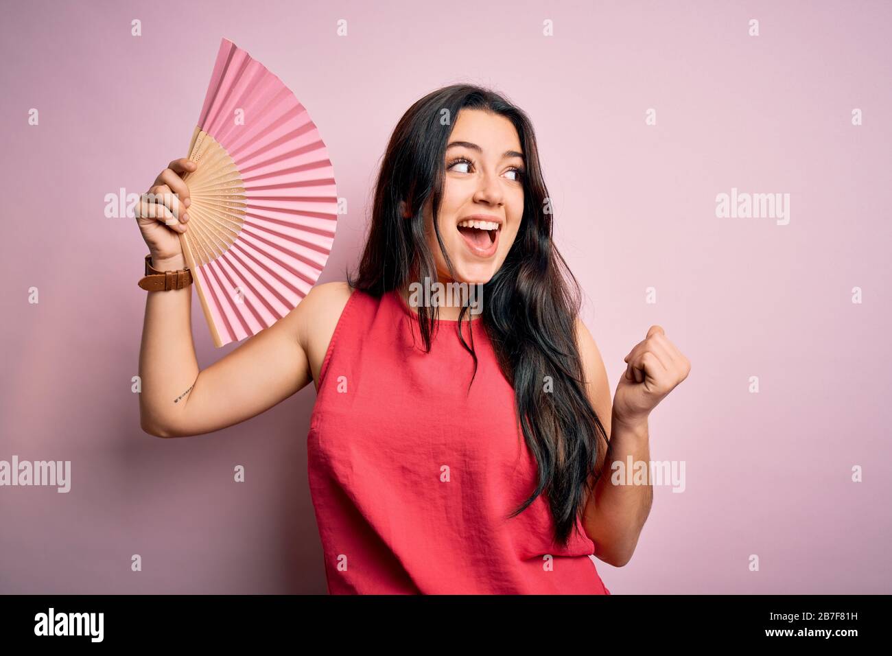 Young brunette woman holding hand fan for fresh air over pink isolated ...