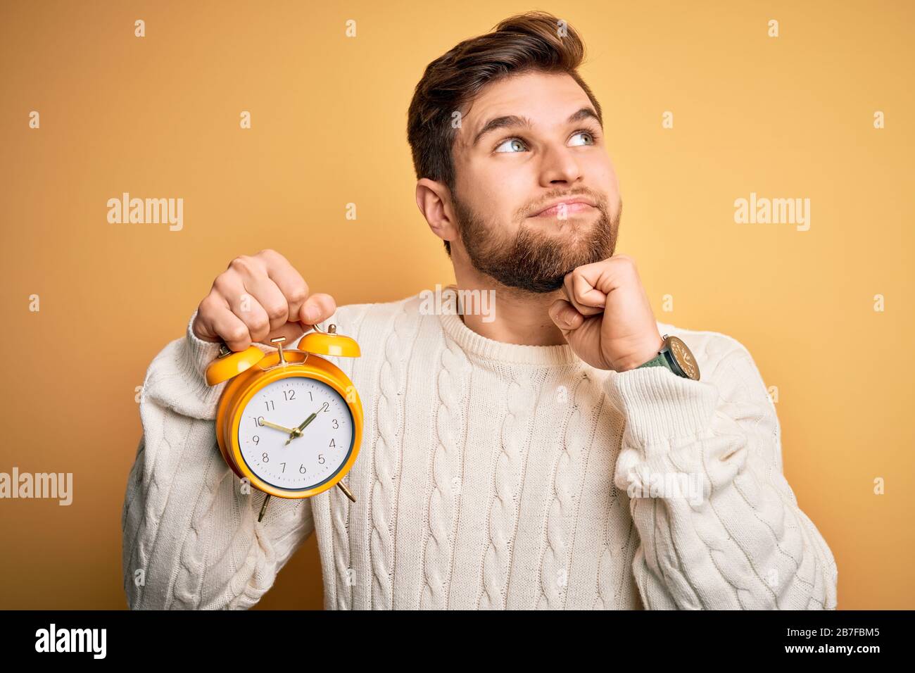 Young blond man with beard and blue eyes holding alarm clock over ...