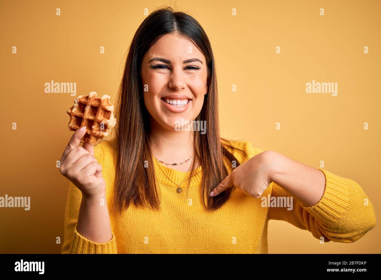 Young beautiful woman eating sweet waffle pastry over yellow background ...
