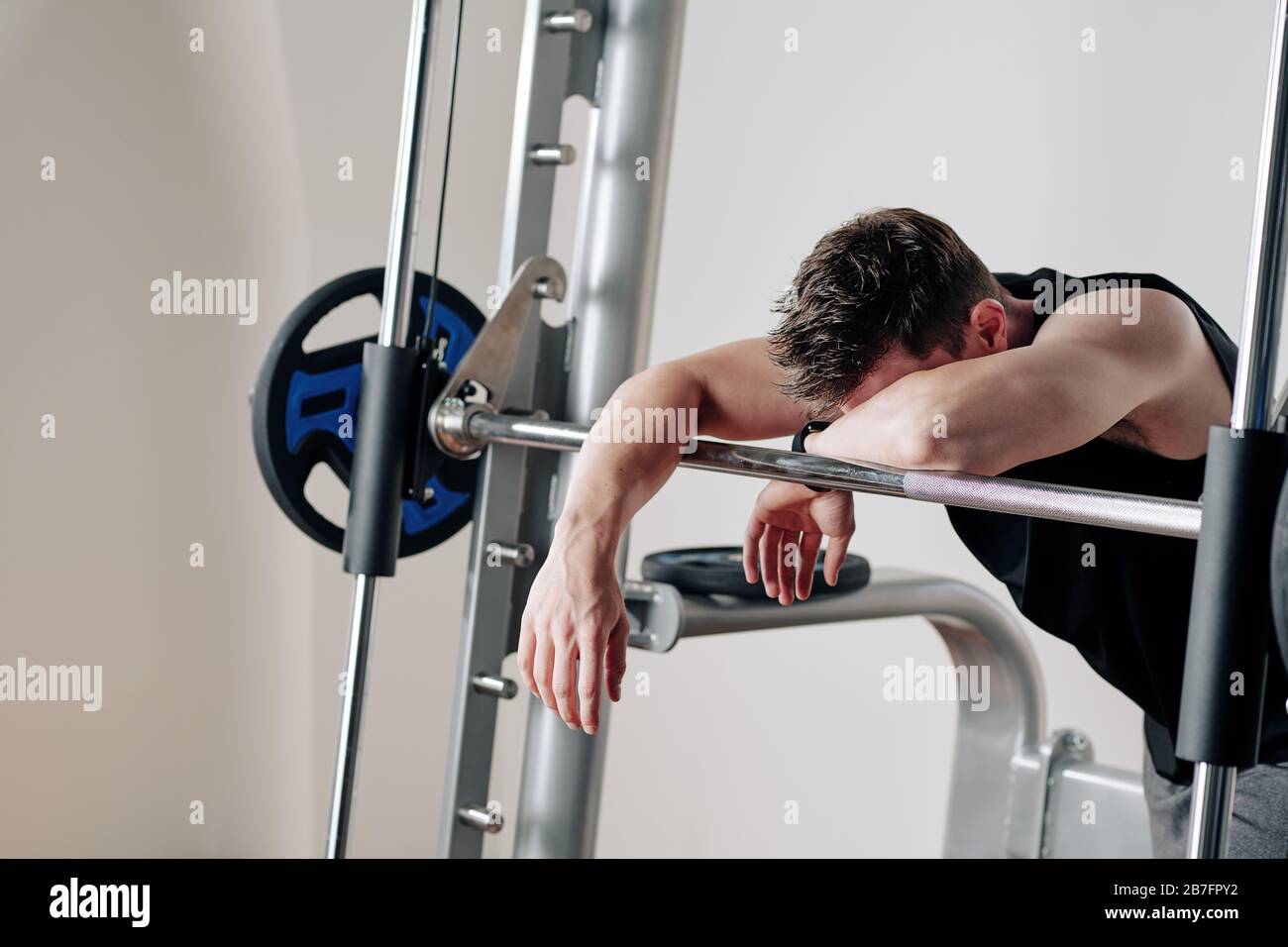 Tired young bodybuilder leaning on barbell after lifting heavy weights ...