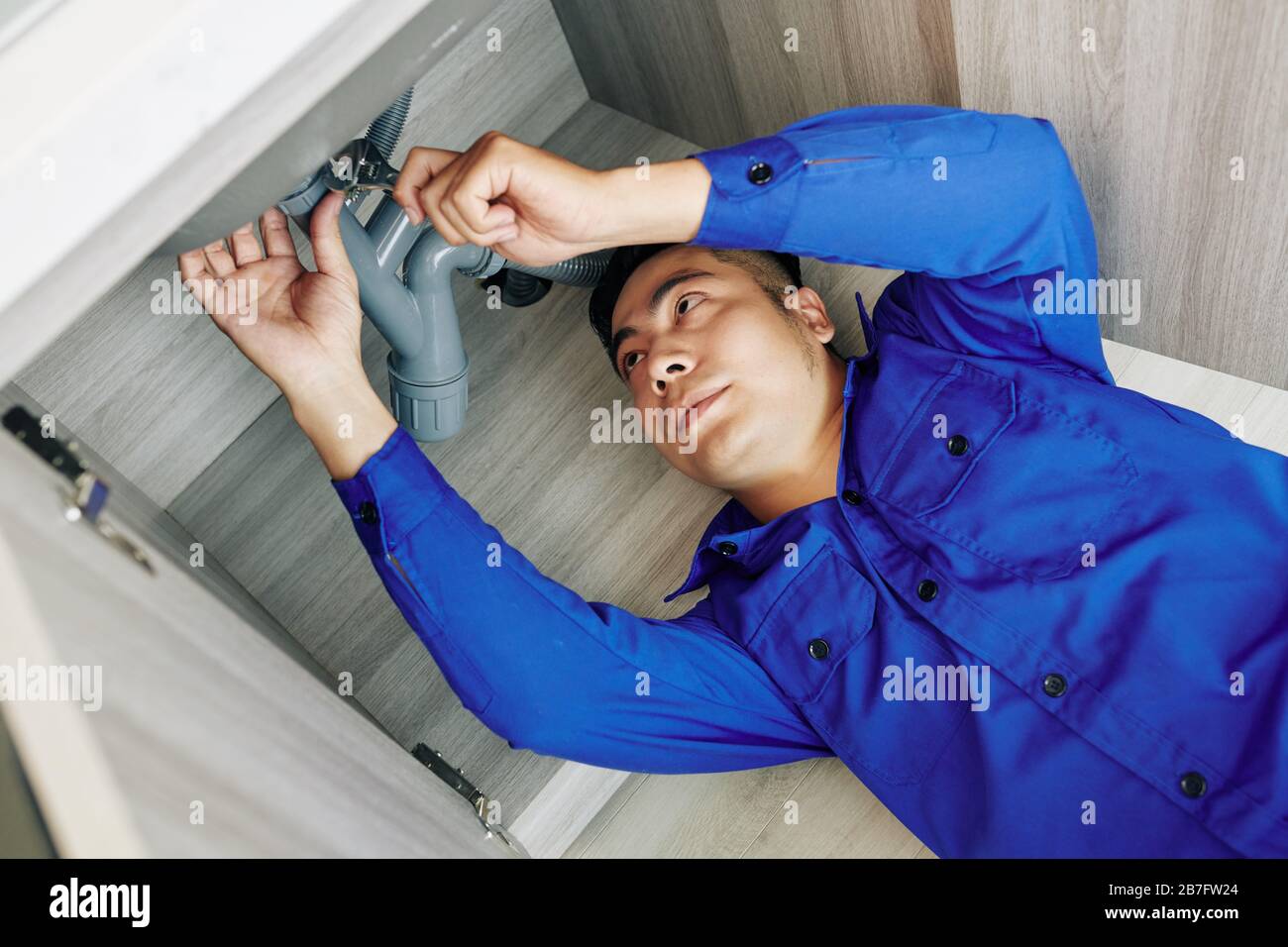 Handome young Asian plumber installing new pipes under kitchen sink ...