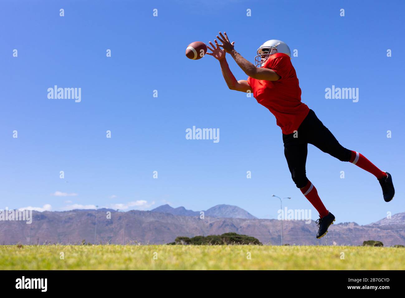 Mixed race male American football player jumping in the air to catch a ...