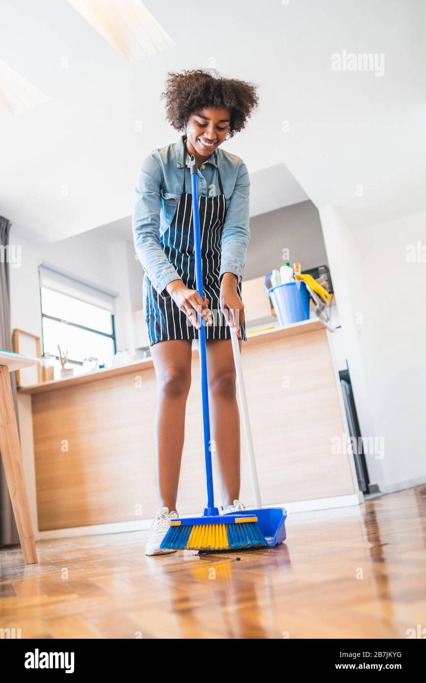 Portrait of young afro woman sweeping wooden floor with broom at home. Cleaning, housework and ...