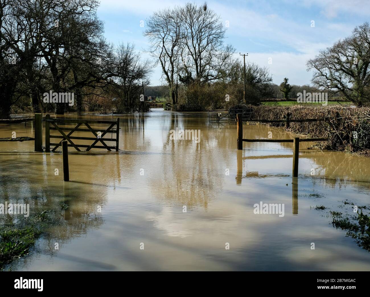 A flooded farm road in winter with partly submerged gate and trees ...