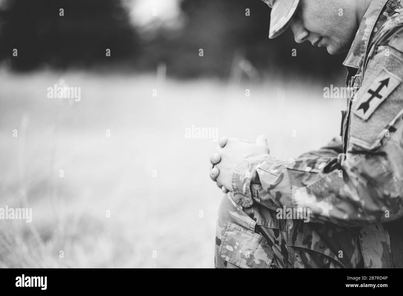 Grayscale shot of a young soldier praying while kneeling on a dry grass ...