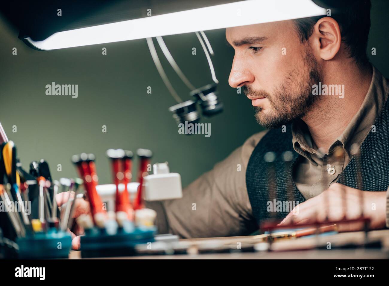 Selective focus of handsome watchmaker working with equipment at table ...