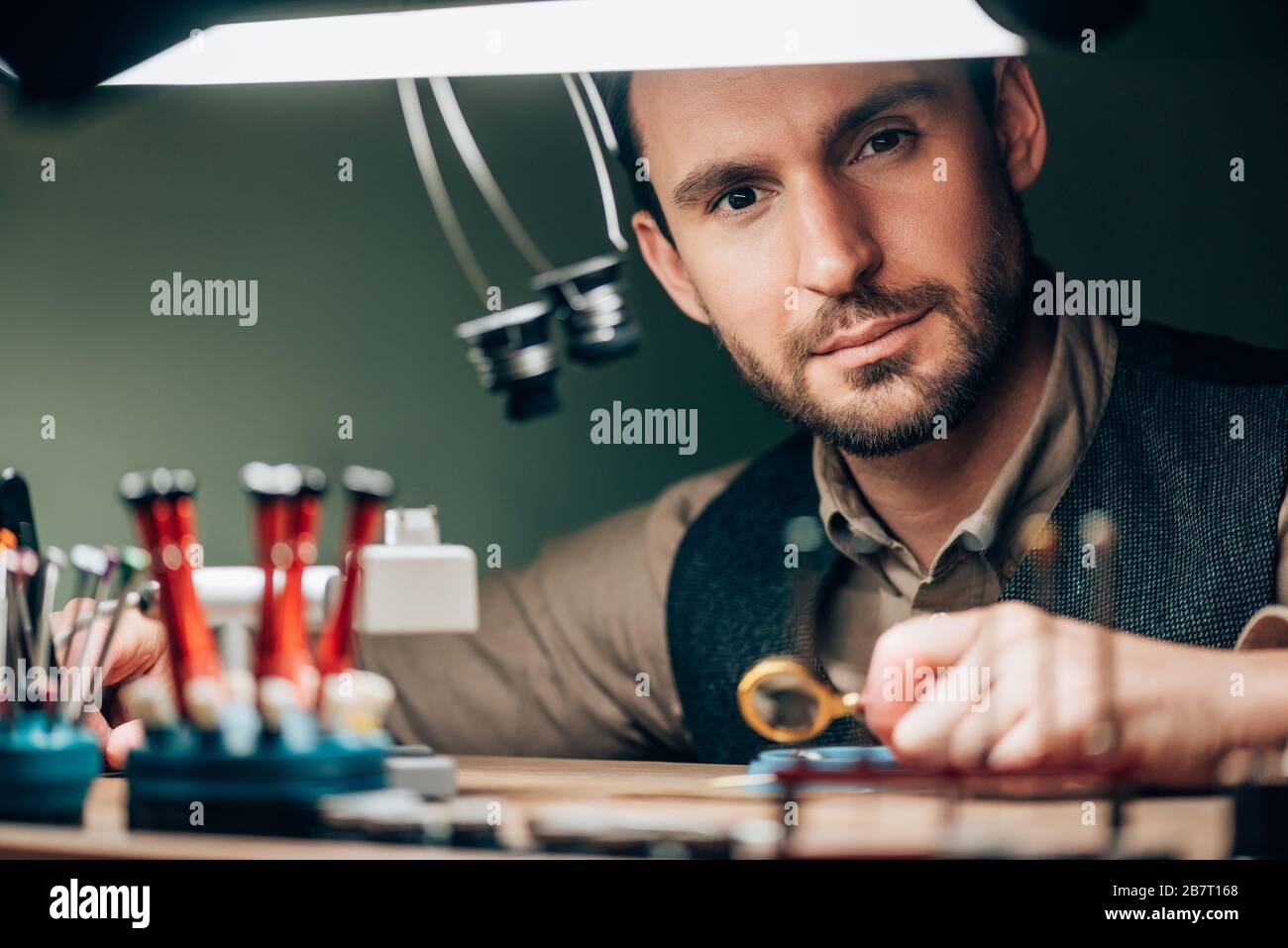 Handsome watchmaker looking at camera while working with watch parts ...
