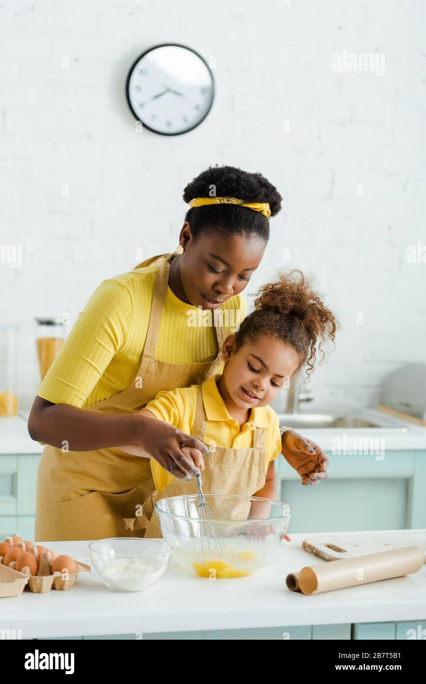 cute african american kid and happy mother holding whisk near bowl while cooking in kitchen ...