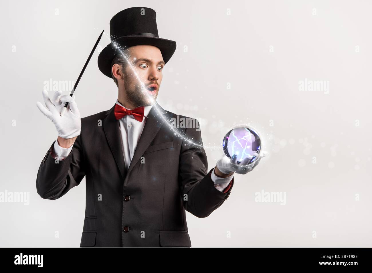 shocked magician holding wand and magic ball isolated on grey with ...