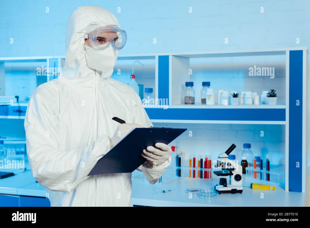 scientist in hazmat suit and goggles holding clipboard and pen near ...
