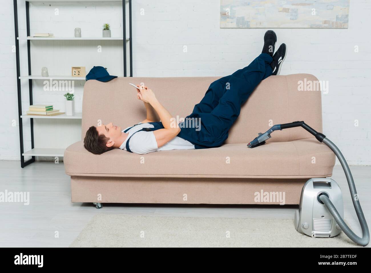 happy cleaner using smartphone while resting on sofa near vacuum ...