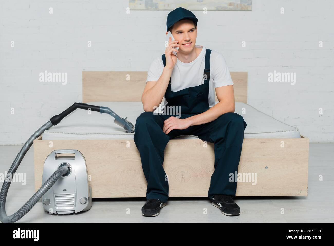 happy cleaner in cap sitting on bed and talking on smartphone near ...
