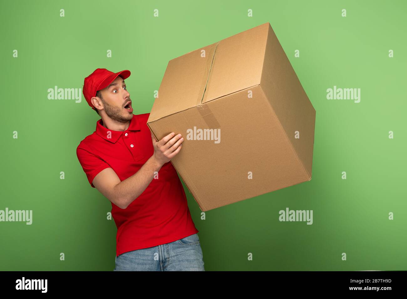 shocked delivery man in red uniform holding huge parcel on green Stock ...