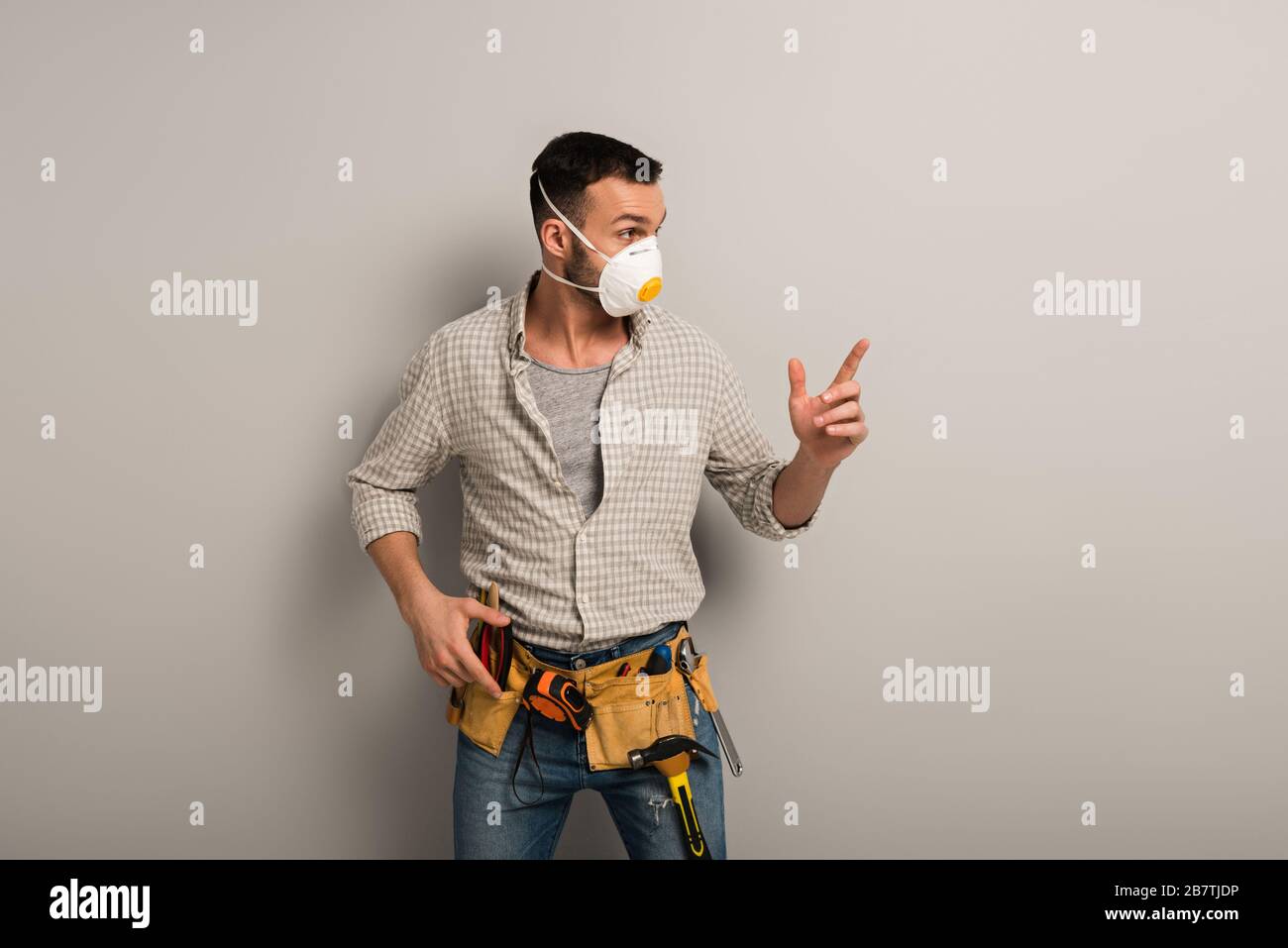 shocked manual worker in safety mask with tool belt pointing on grey ...