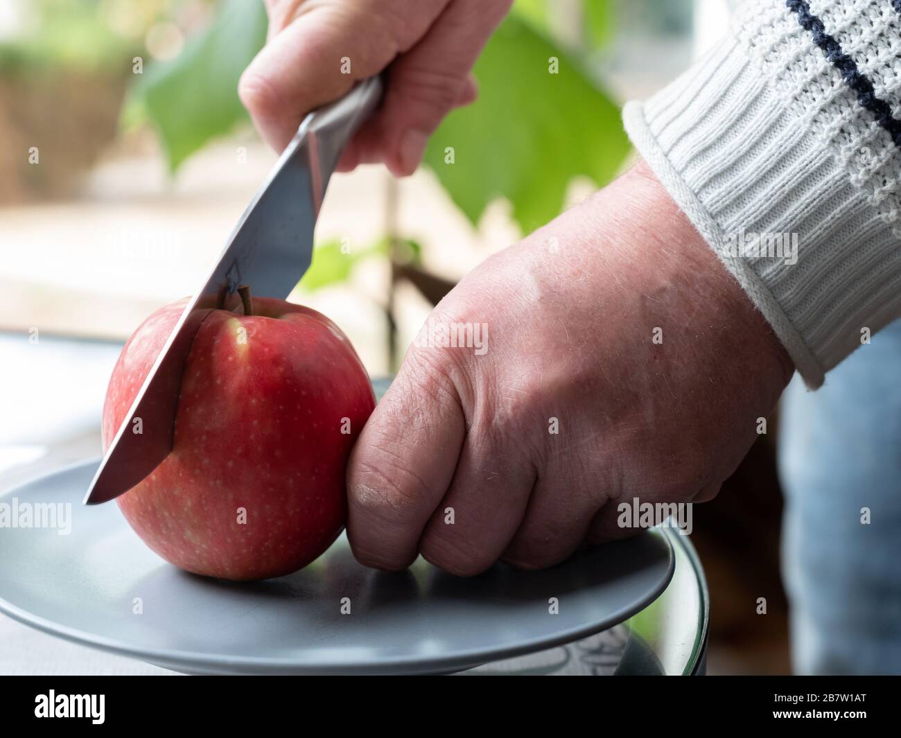 A mans hand with a kitchen knife cuts a ripe red apple on a grey plate ...