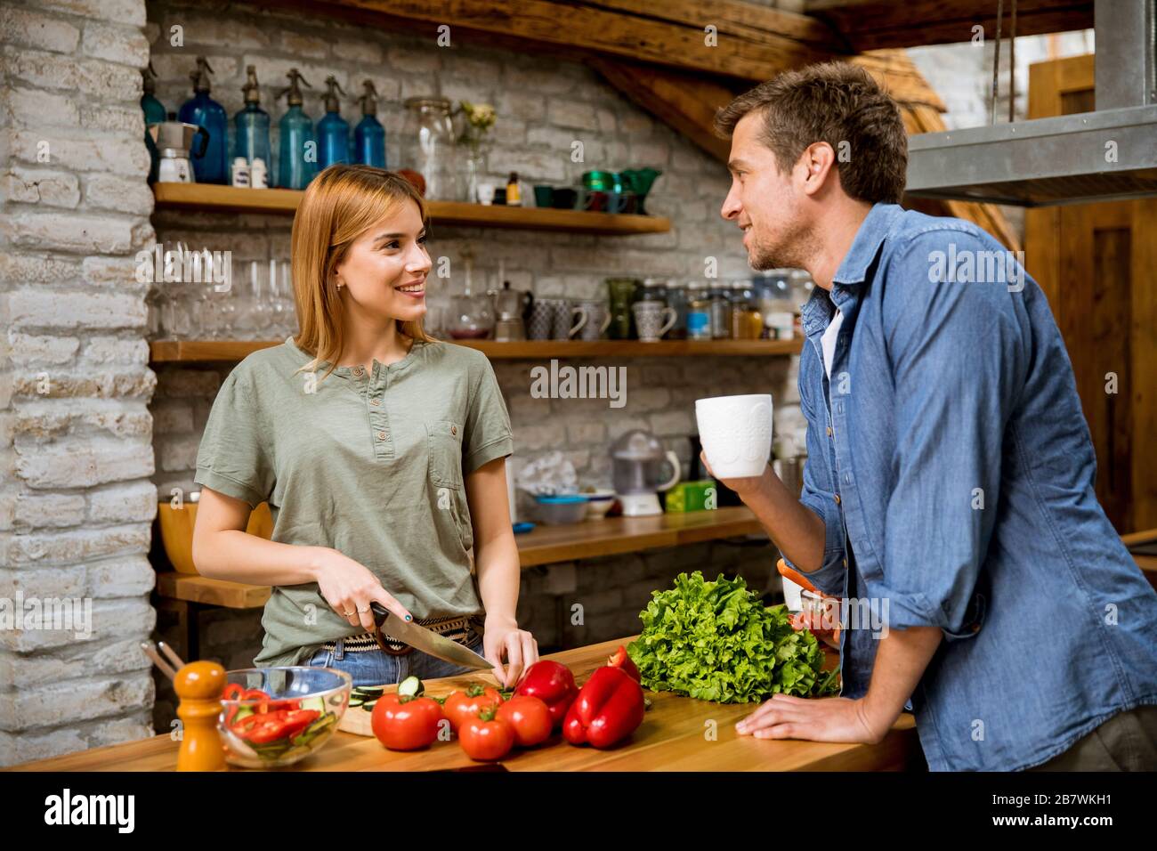 Young woman cooking while man drinking coffee in the rustic kitchen ...