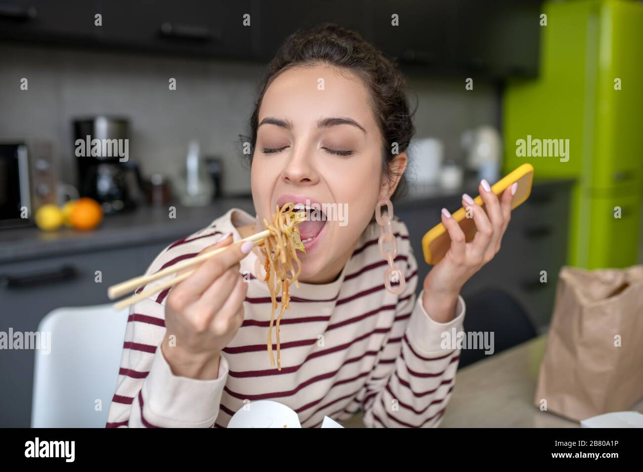 Young woman enjoying noodles in the kitchen, holding mobile Stock Photo ...