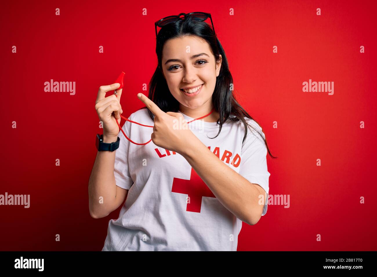 Young lifeguard woman wearing guard equipement holding whistle over red ...