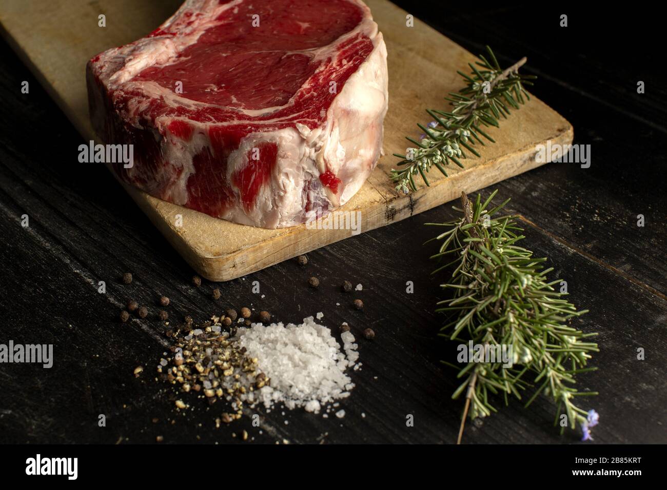 A raw beef chop on a chopping board with herbs and seasoning on a dark ...
