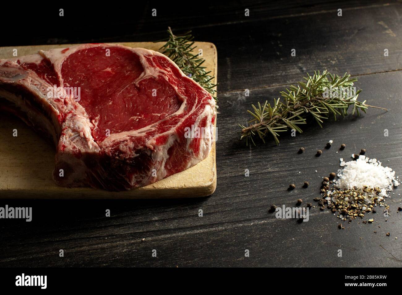 A raw beef chop on a chopping board with herbs and seasoning on a dark ...