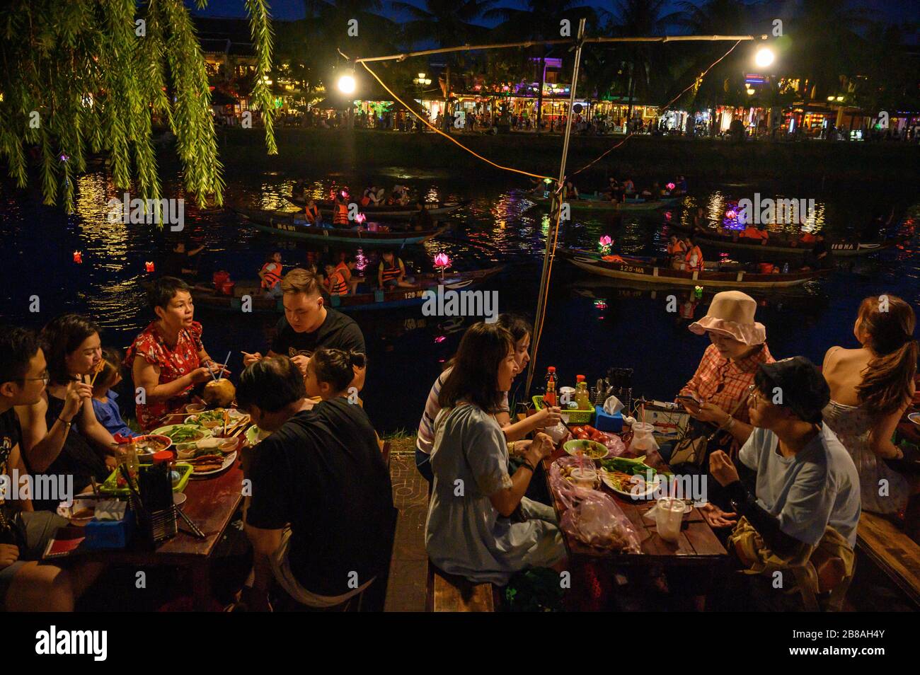 People eating dinner by the water as others row by in boats, Hoi An ...