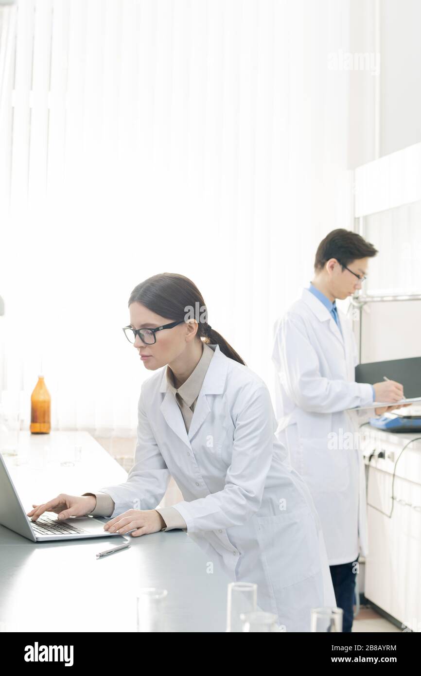 Pretty young female lab worker in whitecoat and eyeglasses standing by ...