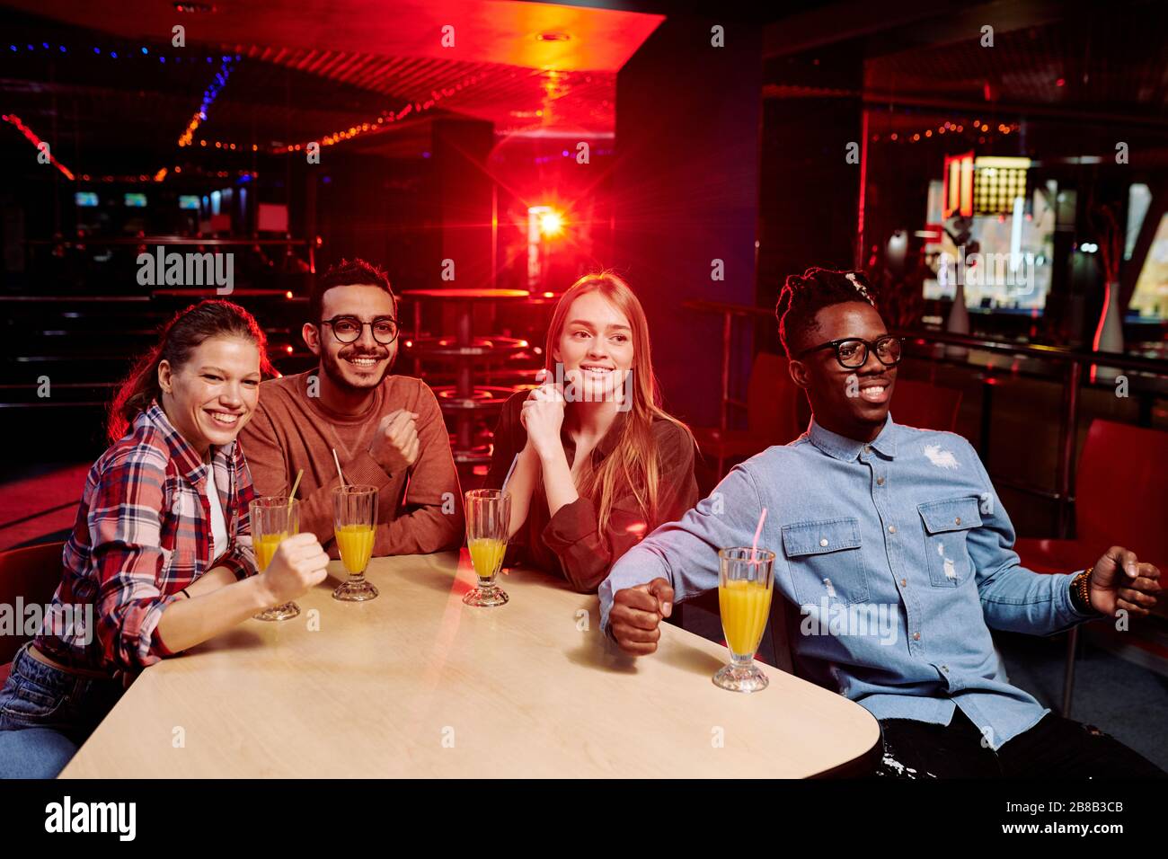 Four young happy intercultural bowling fans sitting by table while ...