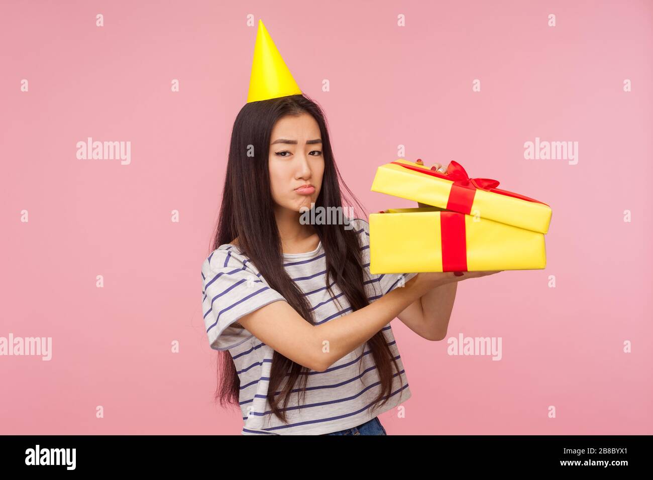 Portrait of unhappy asian girl wearing party cone unpacking gift box ...