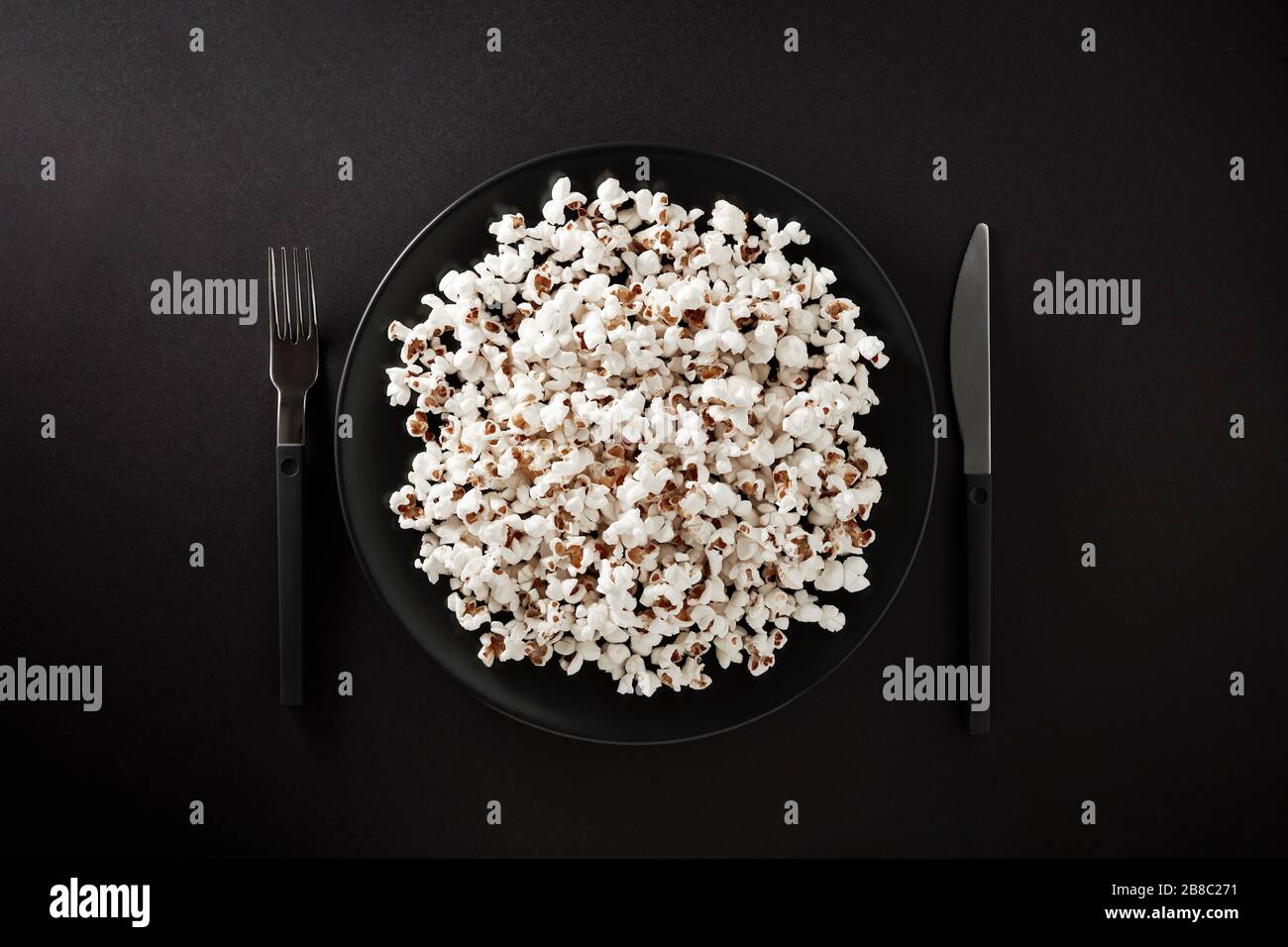 Black plate full of popcorn with fork and knife on black background ...
