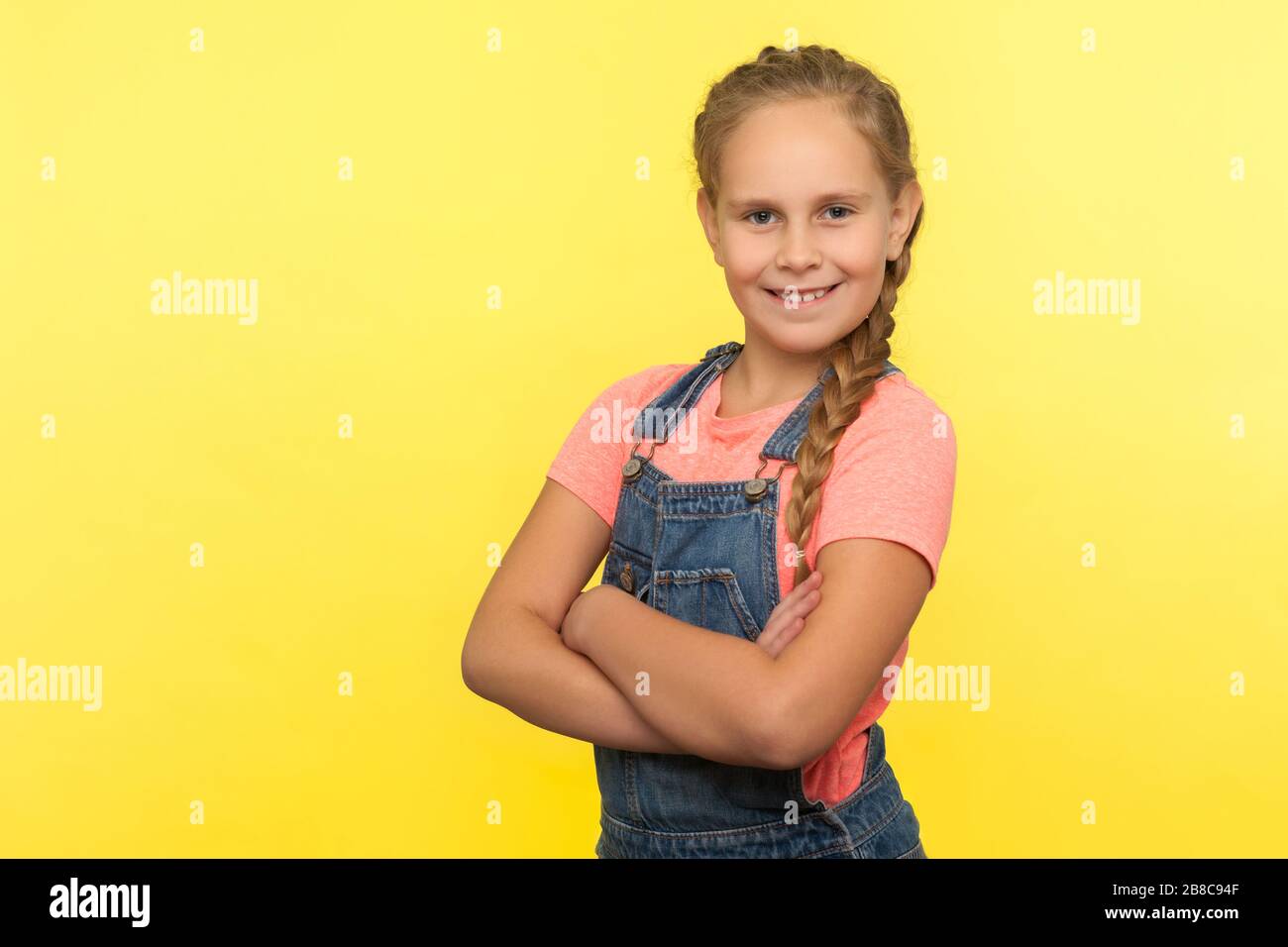 Portrait of cheerful little girl with braid in denim overalls holding ...