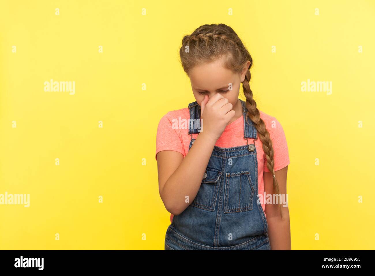Portrait of unhappy little girl with braid in denim overalls hiding ...