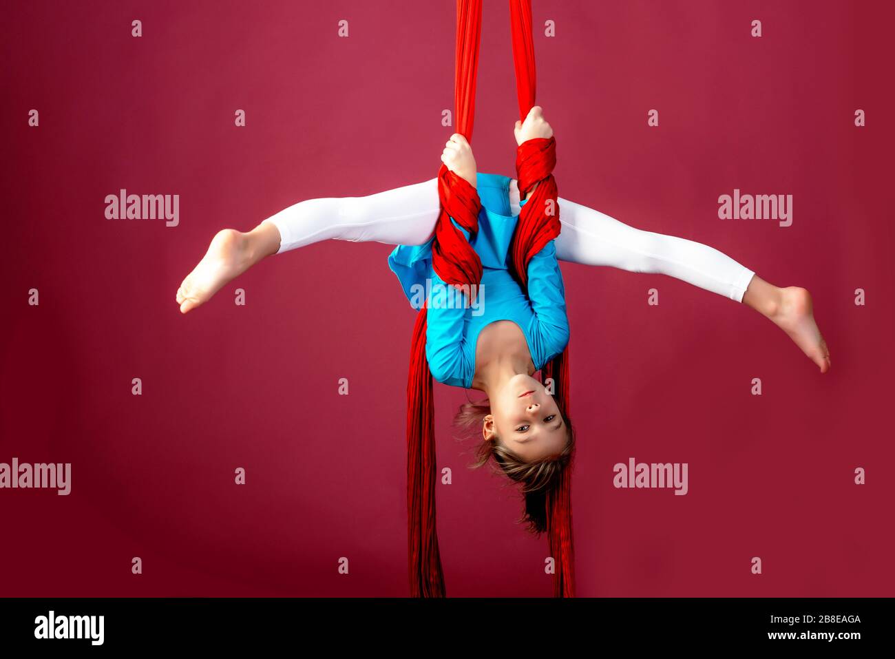 Little acrobat girl doing splits in the air holding on to a red air ...