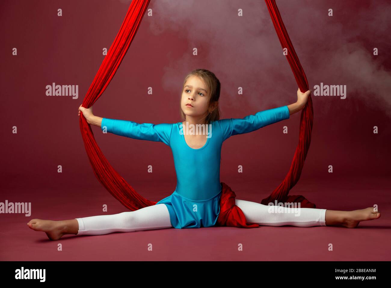 Little acrobat girl doing splits in the air holding on to a red air ...